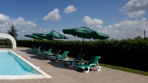 Piscina retangular com deck branco, rodeada por cadeiras de grama e guarda-sóis, sob um céu claro.