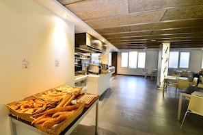 Uma área de buffet com pães e doces, uma cozinha ao fundo e uma sala de jantar com mesas e cadeiras.