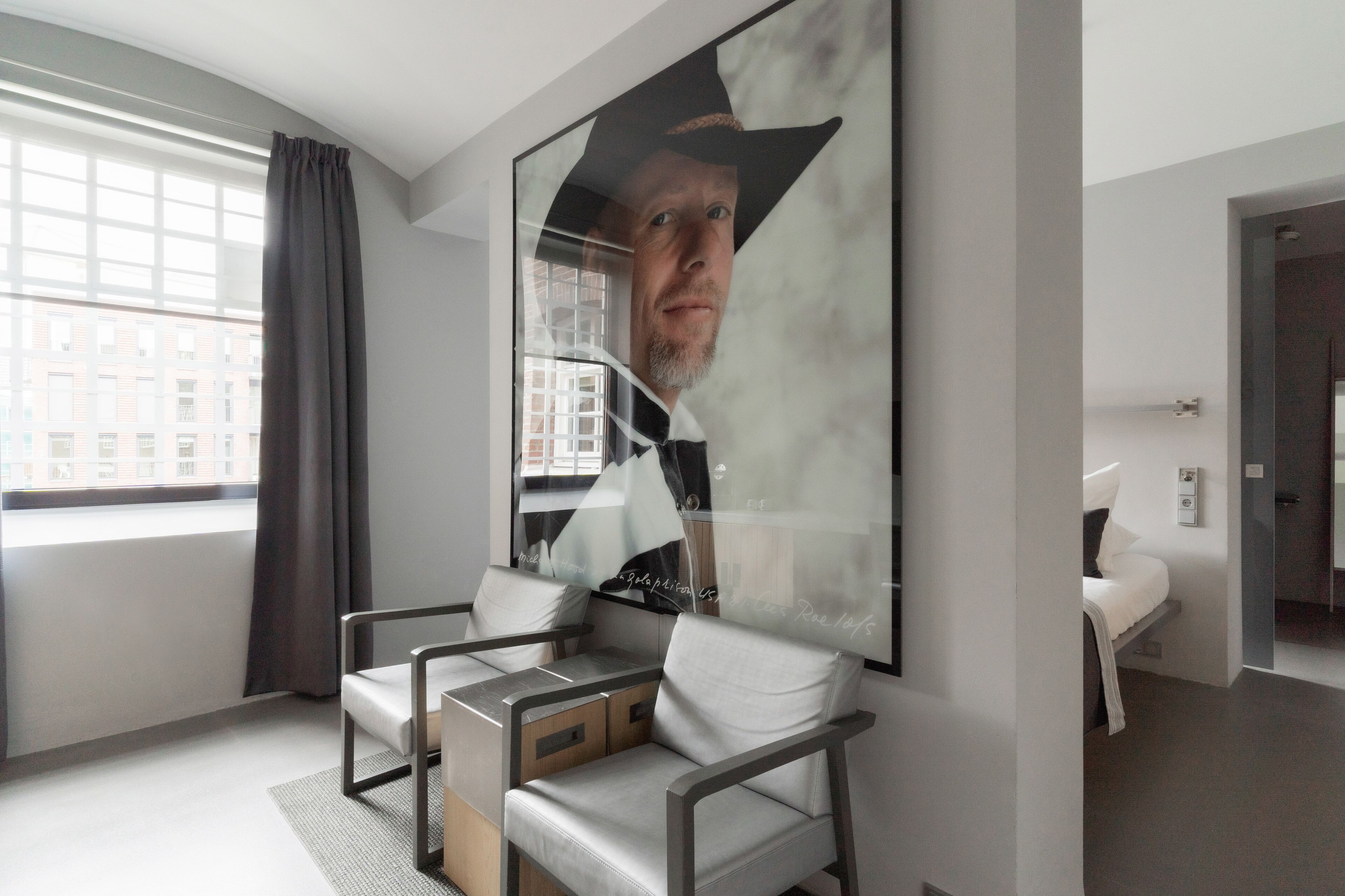 Quarto de hotel moderno com uma janela grande, uma cama, área de estar com uma cadeira e uma pequena mesa, e uma grande obra de arte abstrata na parede.