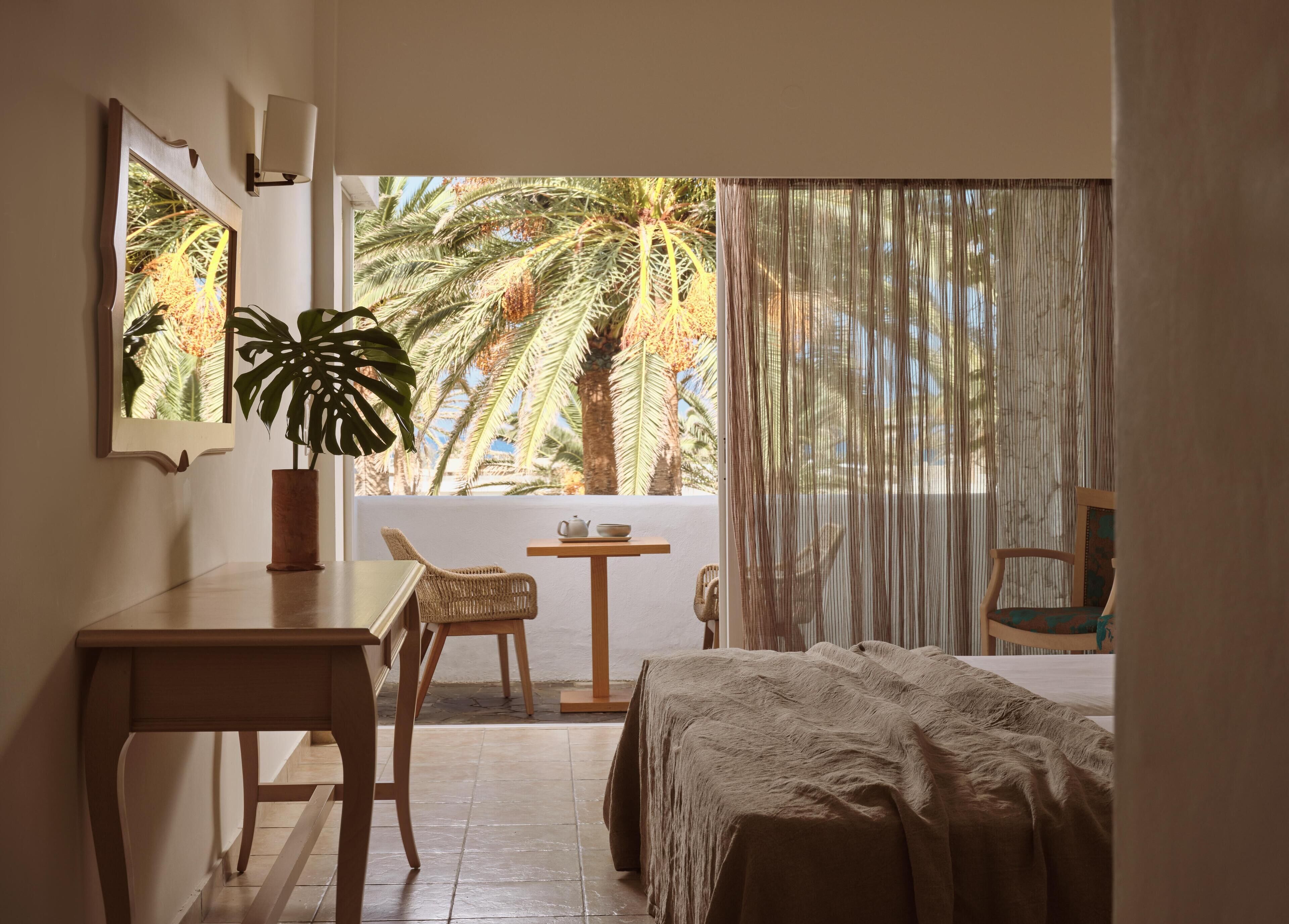 Quarto com cama, escrivaninha, cadeira, planta em vaso e vista para palmeiras através de cortinas translúcidas.
