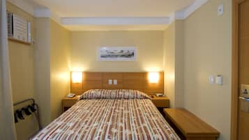 Quarto de hotel com cama, mesas de cabeceira, um banco de madeira, um quadro pendurado e um aquecedor fixado na parede.