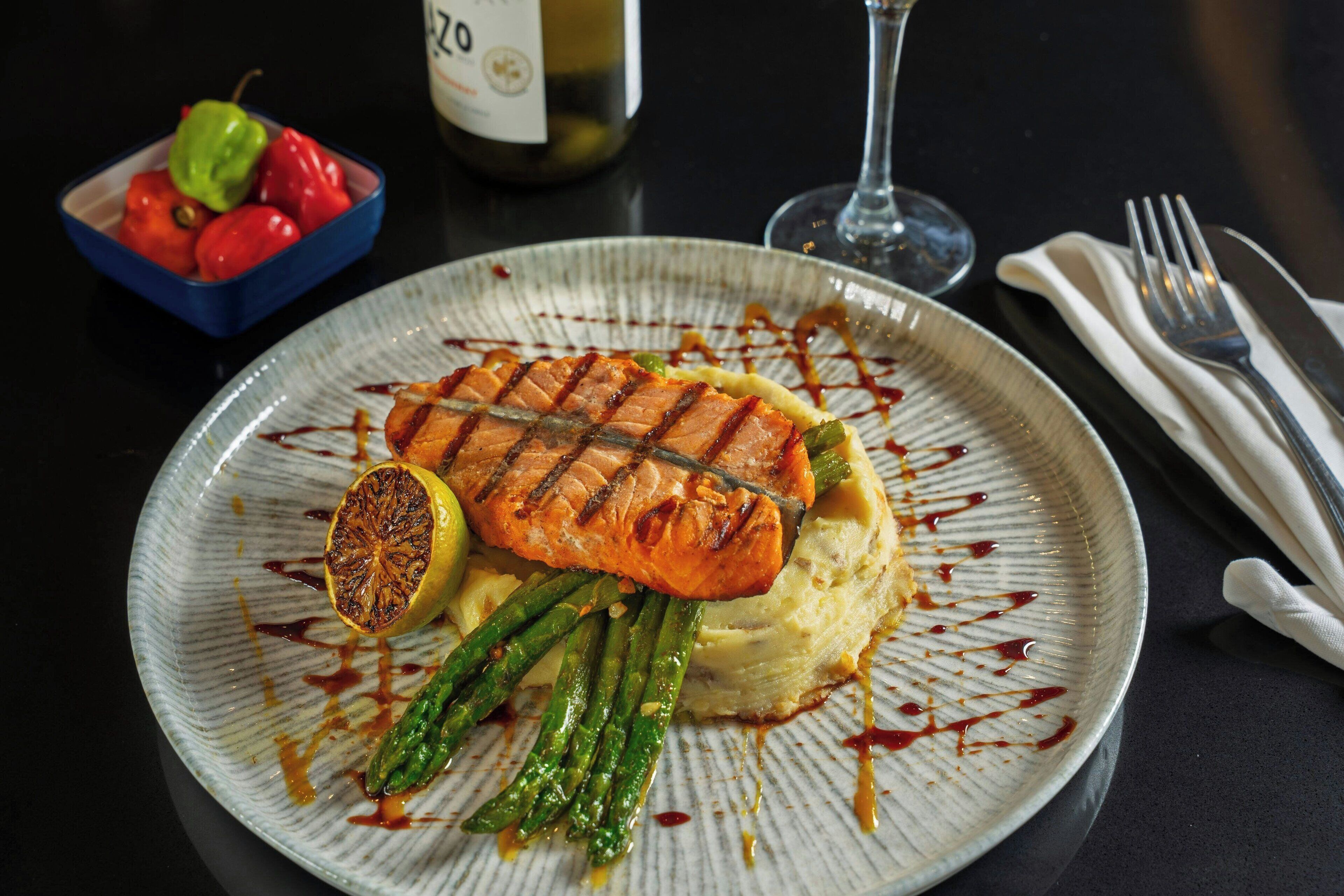 Prato com salmão grelhado, aspargos e uma fatia de limão, acompanhado de uma garrafa de vinho e um copo.