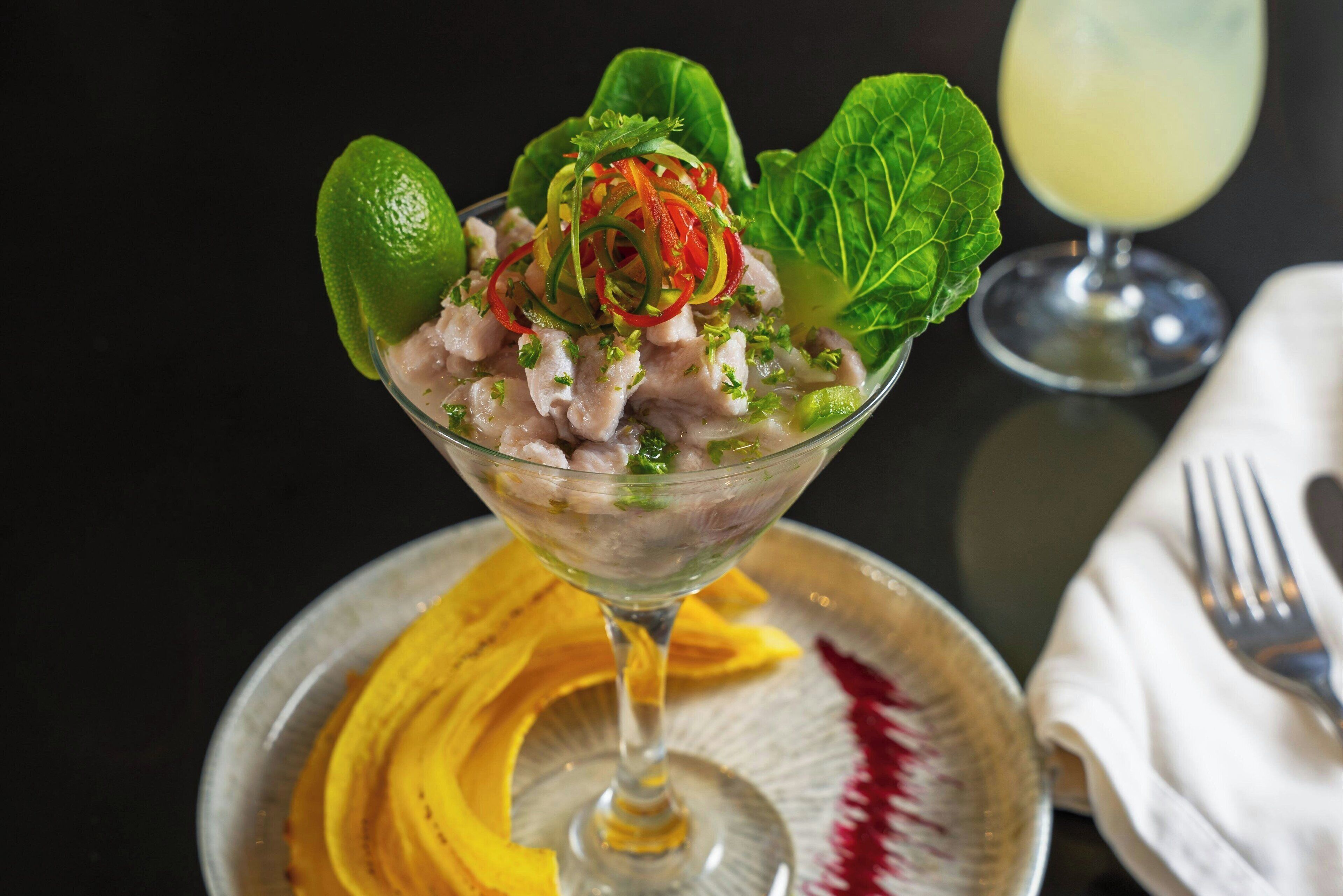 Um copo de coquetel de frutos do mar, decorado com limão e ervas, servido em um prato com padrão amarelo e vermelho, acompanhado por um copo de cocktail e um garfo sobre uma folha de papel.