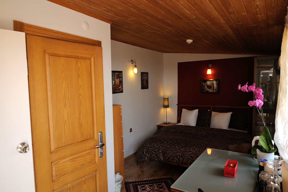 Quarto de hotel com porta de madeira, cama com colcha de padrão de zebra, mesa de cabeceira com abajur, vaso com orquídeas cor-de-rosa e mesa com vela e bandeja de copos.