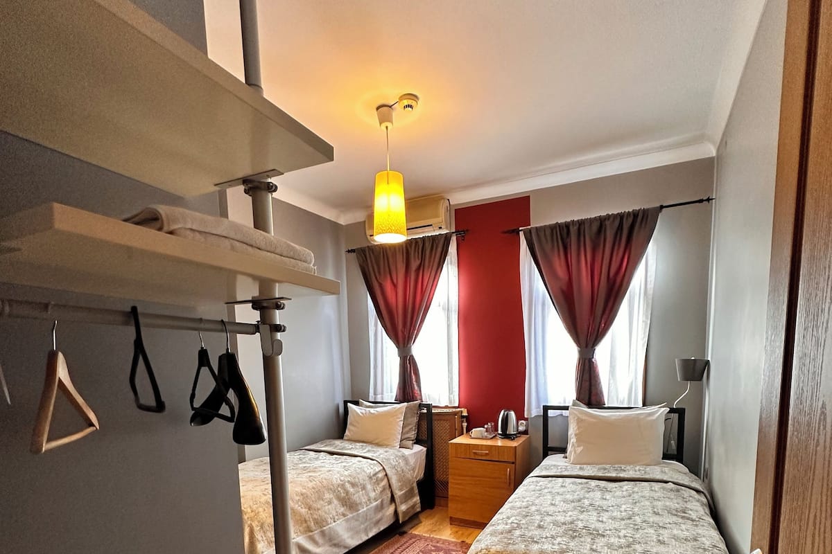 Quarto de hotel com duas camas, piso de madeira, parede de destaque vermelha e luminária pendente.