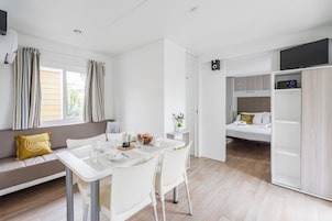 Quarto moderno e bem iluminado, com mesa de jantar, cadeiras, sofá com almofadas, cama e TV fixada na parede.