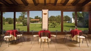 Uma área de refeições com mesas preparadas para quatro pessoas, grandes janelas com vista para um jardim paisagístico.