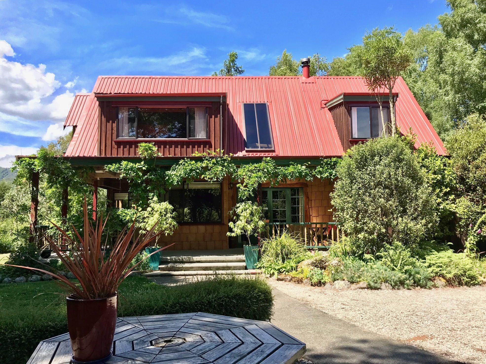 Uma casa de dois andares com telhado vermelho, revestimento de madeira e varanda frontal com corrimão verde.