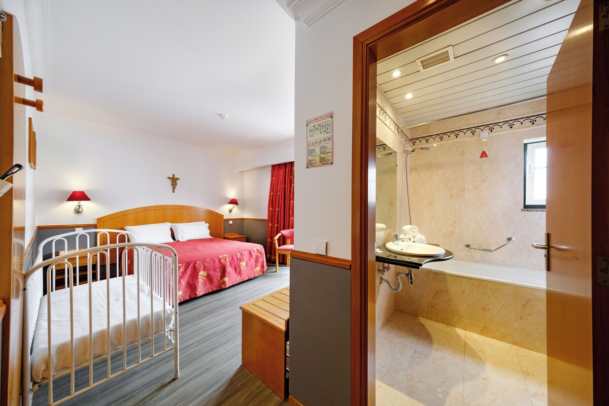 Quarto de hotel com cama, escrivaninha, cadeira, mesinha, banheiro com pia, espelho e chuveiro.