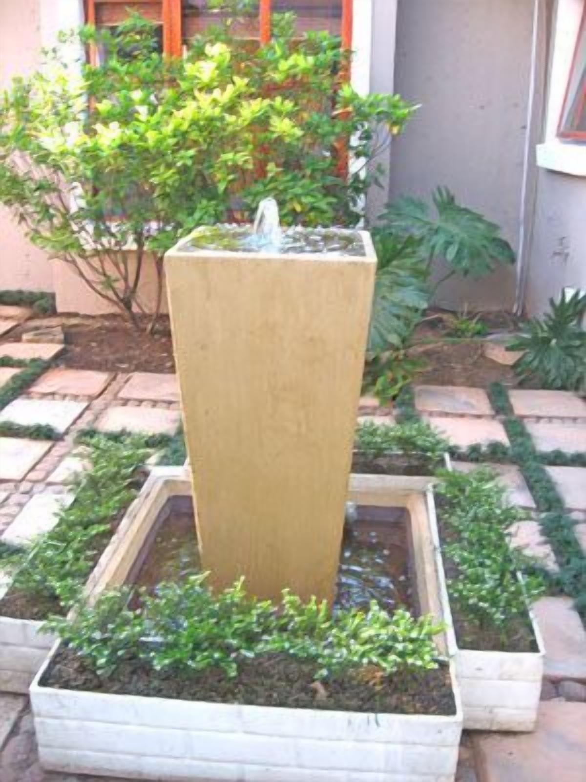 Um espelho d'água retangular com um bocal central, rodeado por um canteiro quadrado com plantas verdes.