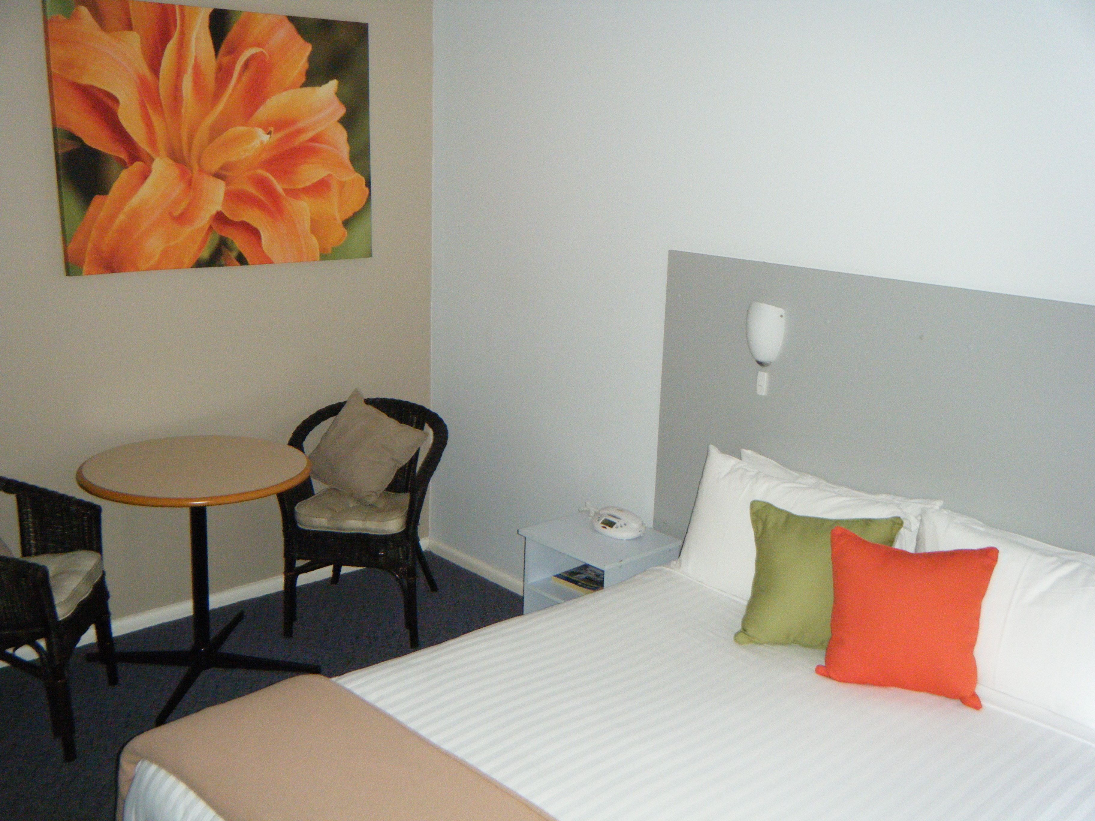 Quarto de hotel com cama, uma cadeira, uma mesinha e uma mesa de cabeceira com abajur.