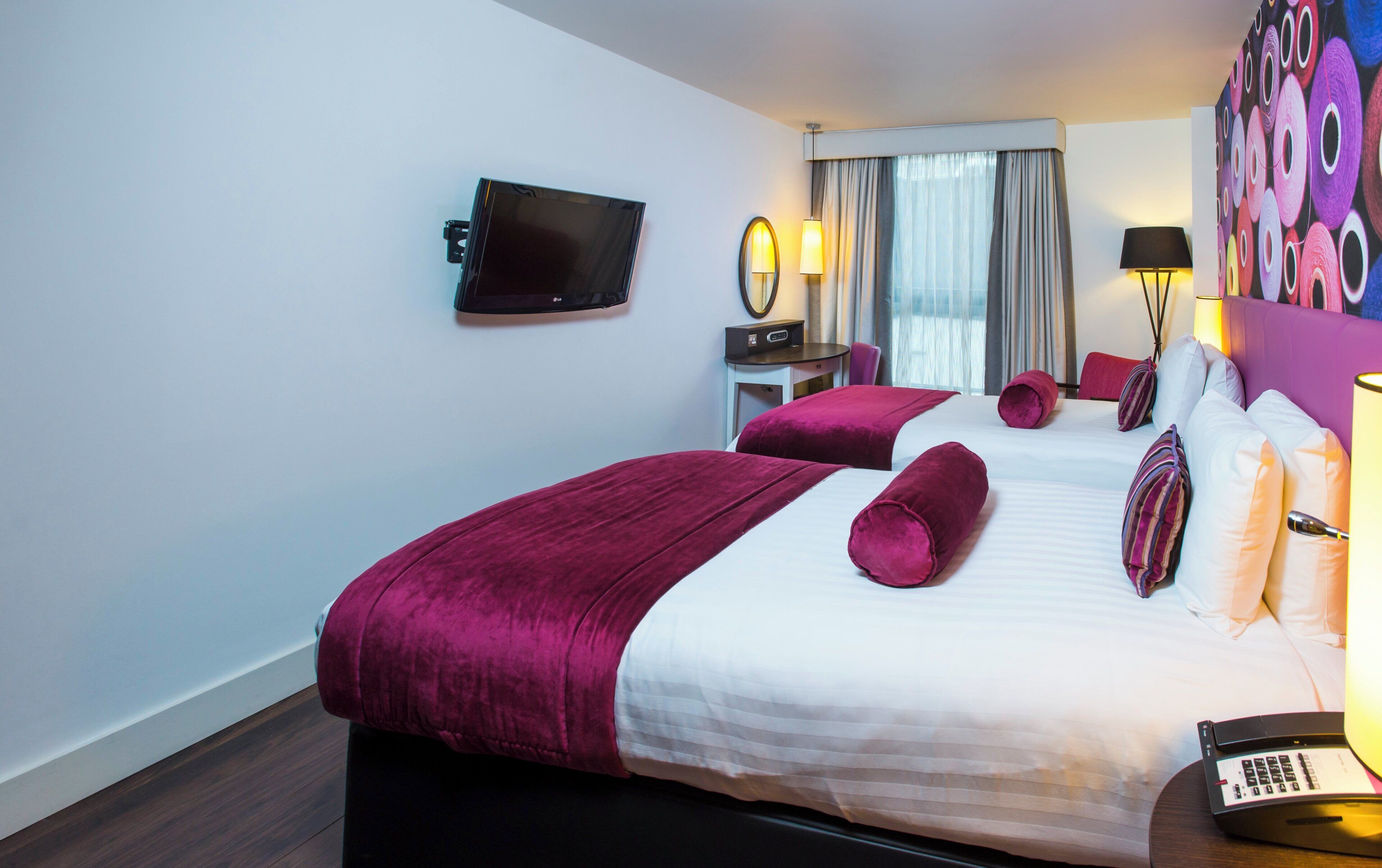 Quarto de hotel com duas camas, televisão de tela plana, escrivaninha com luminária e uma janela grande com cortinas.