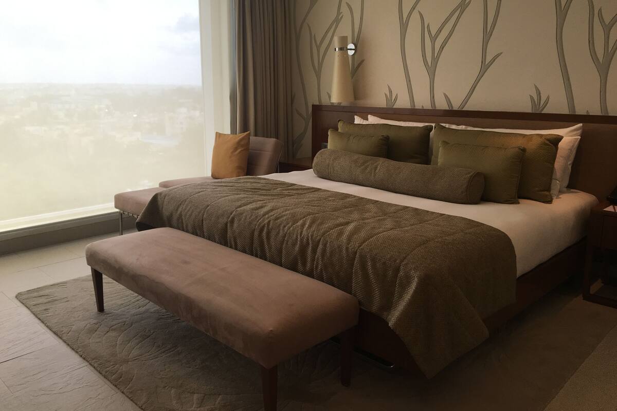 Quarto com cama grande, um banco, uma mesa de cabeceira e vista para a cidade através de uma grande janela.