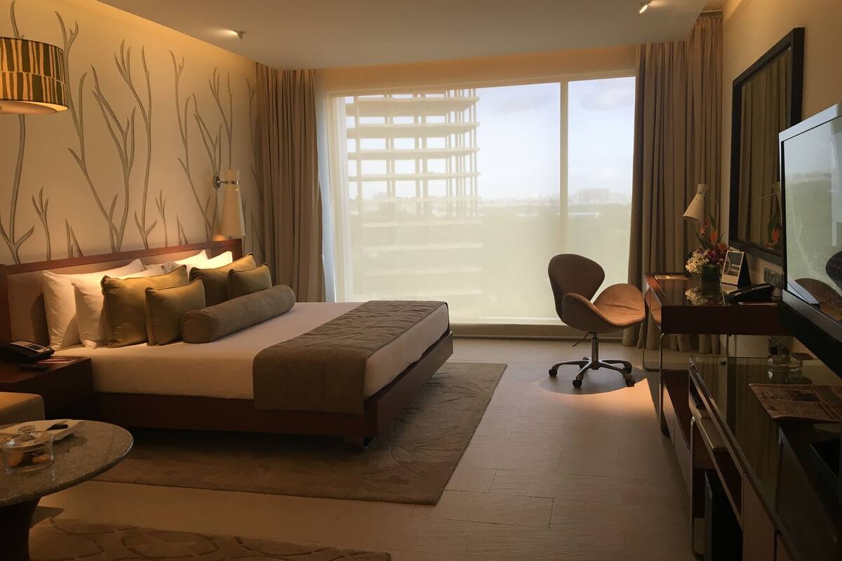 Quarto de hotel moderno com uma cama grande, escrivaninha, televisão e vista da cidade através de amplas janelas.