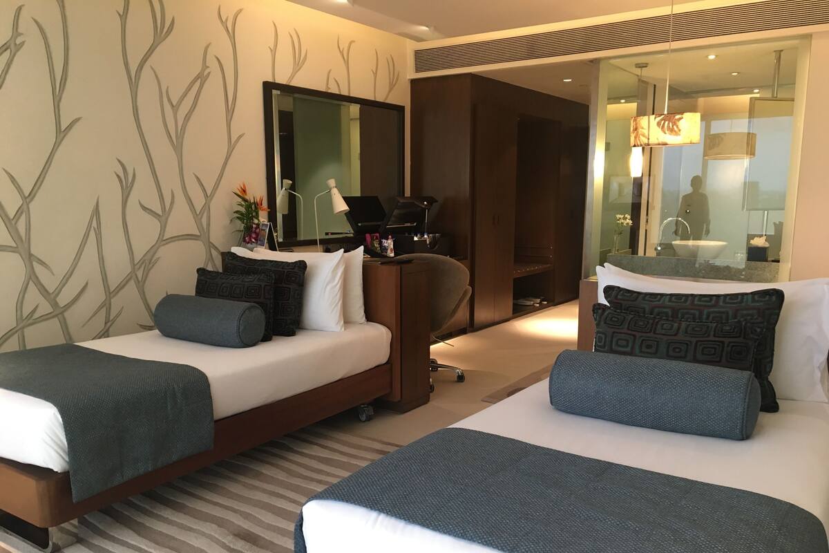 Quarto de hotel com duas camas, uma mesa, uma cadeira, um espelho e um banheiro visível através de uma porta de vidro.