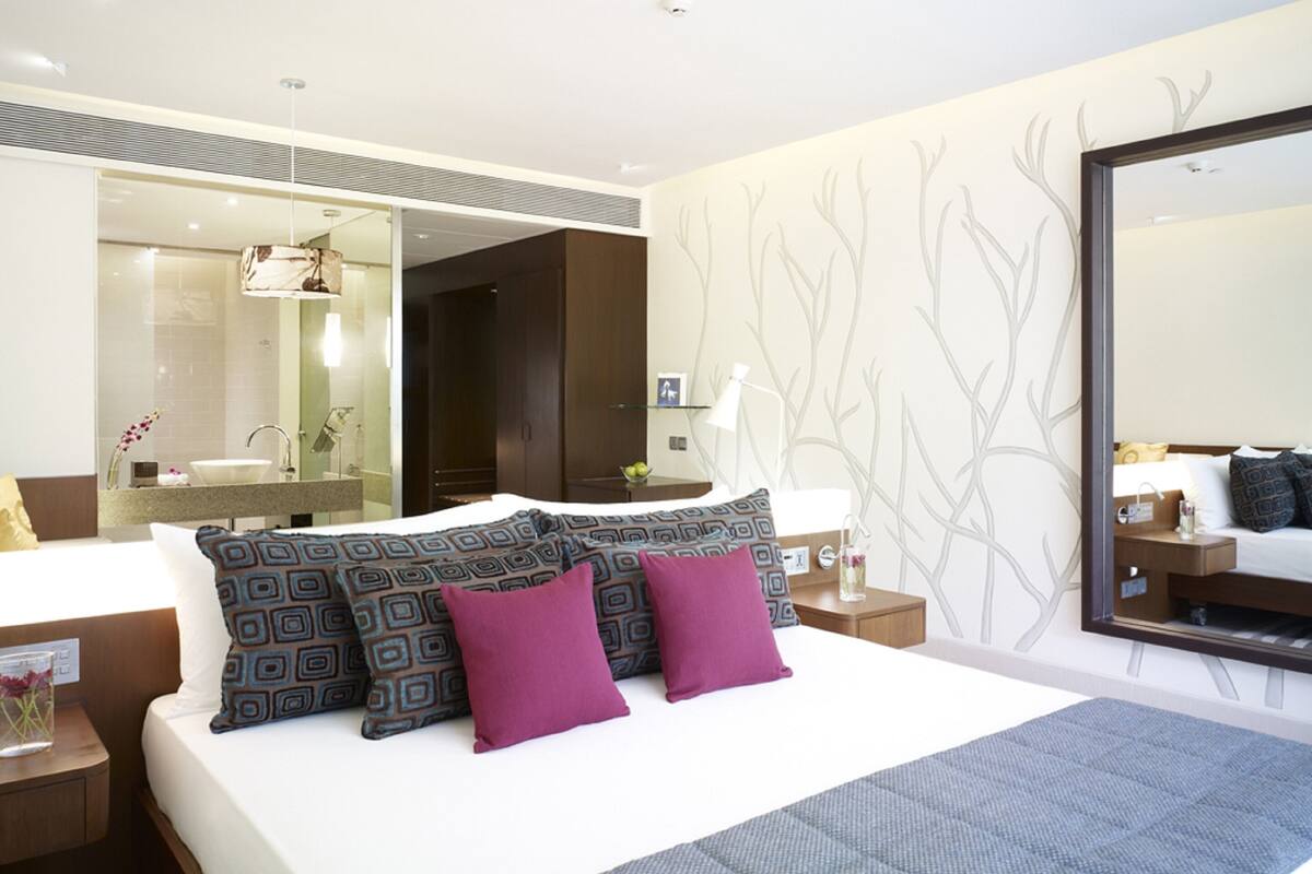 Quarto de hotel moderno com uma cama grande, duas mesas de cabeceira, um criado-mudo, um espelho e um banheiro visível através de uma porta aberta.