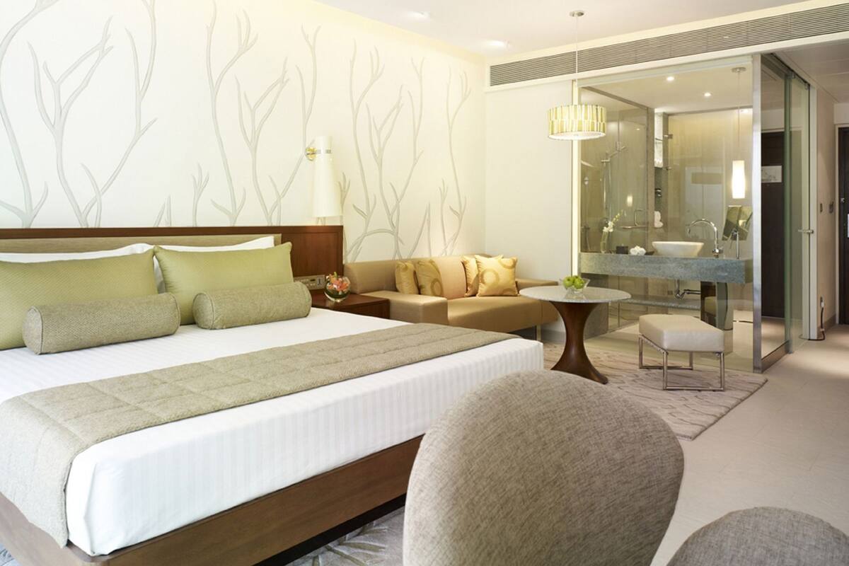 Quarto de hotel moderno com uma cama grande, um sofá, uma pequena mesa redonda e um banheiro com paredes de vidro.