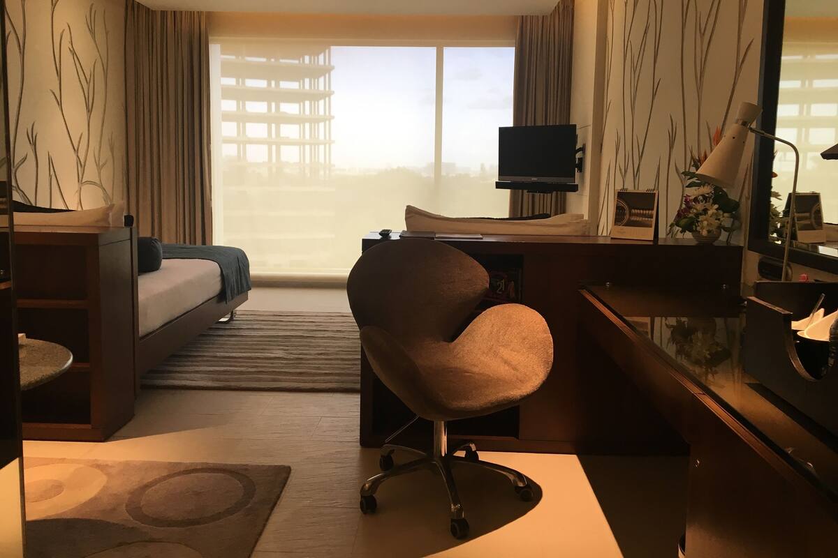 Quarto de hotel com uma cama, uma escrivaninha com um computador, uma cadeira, uma televisão e uma janela grande com vista para um edifício.