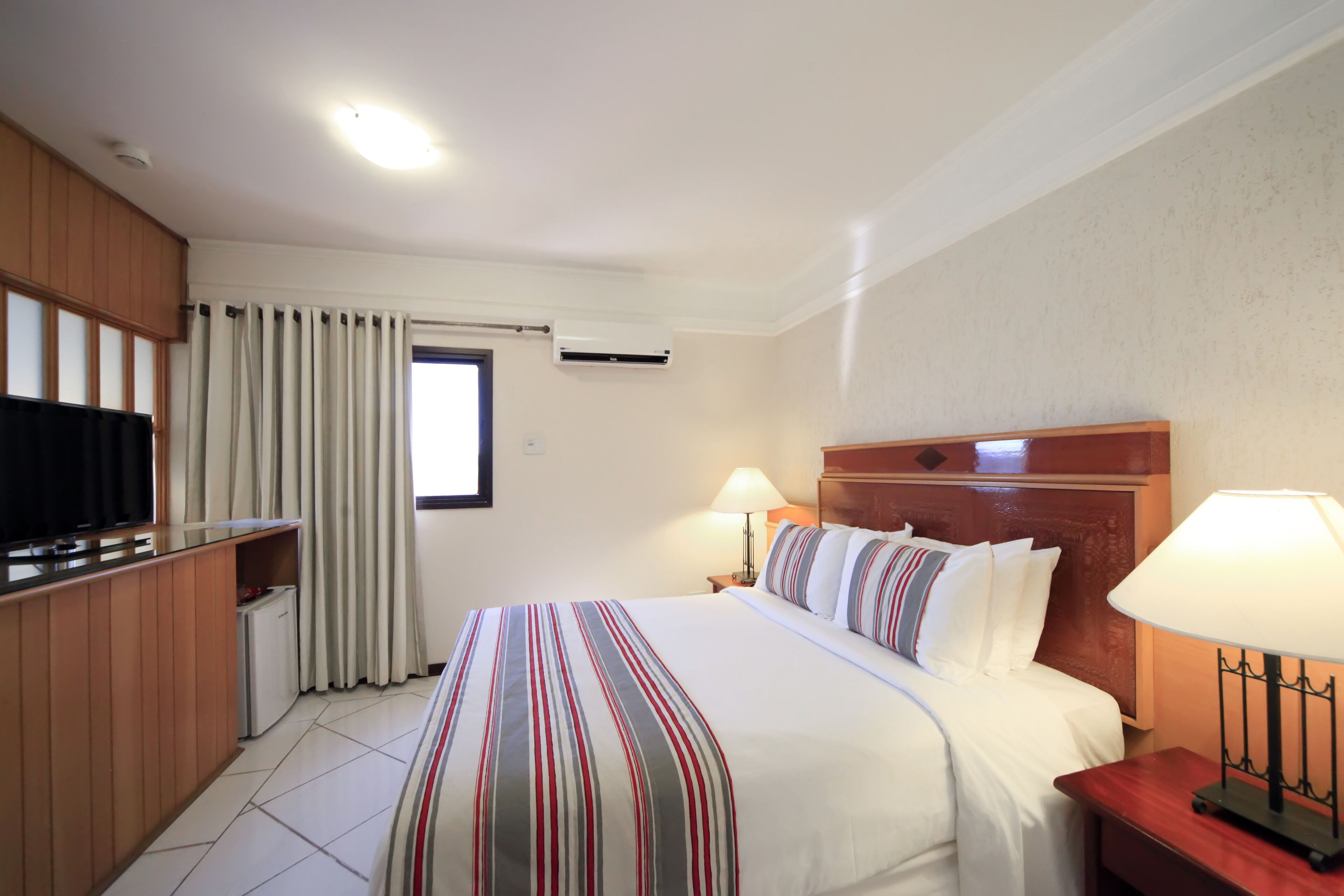 Quarto de hotel com cama, luminárias de cabeceira, TV, um pequeno espaço de cozinha e janela com cortinas.