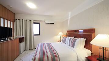 Quarto de hotel com cama, luminárias de cabeceira, TV, um pequeno espaço de cozinha e janela com cortinas.