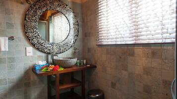 Casa de banho com espelho redondo, pia com produtos de higiene, um cesto de lixo e um banquinho de fibra natural.