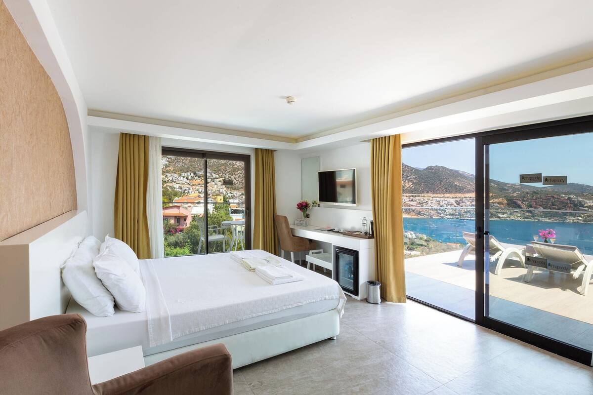 Quarto de hotel com cama, escrivaninha e uma grande porta de vidro deslizante que dá para uma varanda com vista para o mar e as colinas.