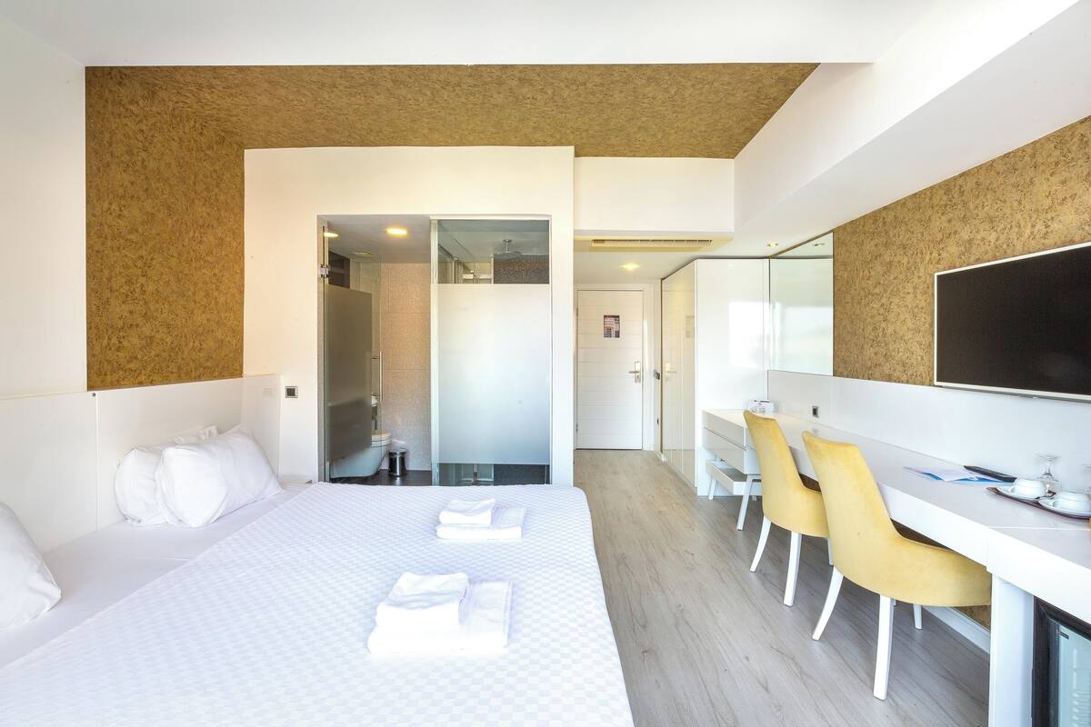 Quarto de hotel moderno com uma cama grande, uma escrivaninha com cadeiras e uma televisão.