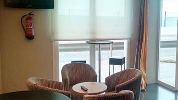 Quarto de hotel com televisão fixada na parede, extintor de incêndio, mesa redonda com duas cadeiras e vista para o mar através de grandes janelas.