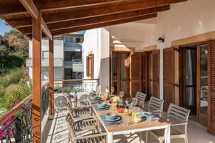 Terraço com mesa de madeira posta para refeições, rodeado de cadeiras, com vista para um edifício e vegetação ao fundo.