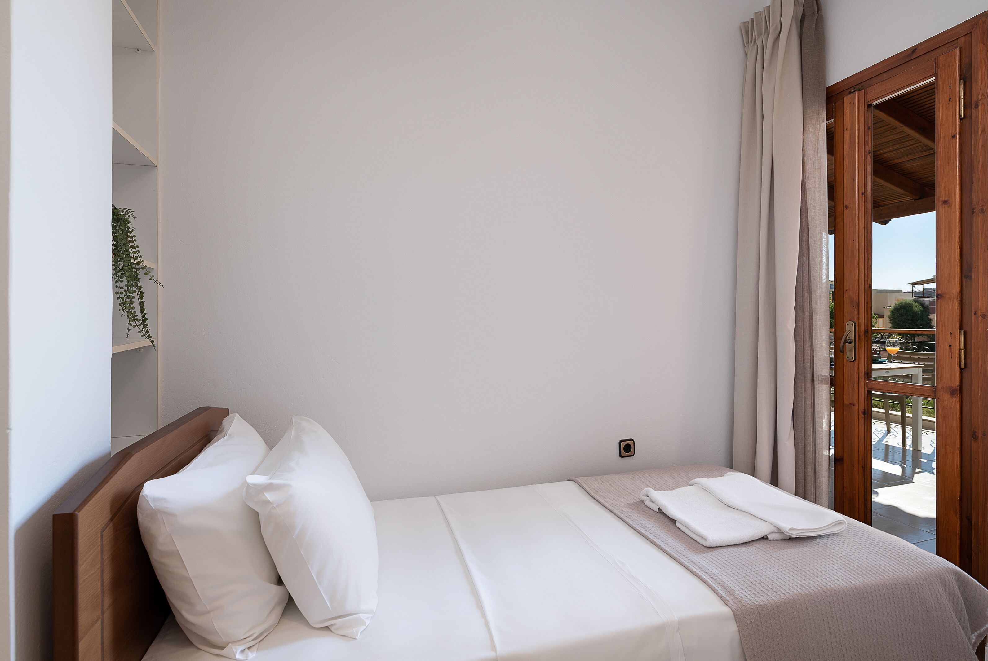 Cama bem arrumada com roupa de cama branca e cabeceira de madeira, uma pequena estante com uma planta, e vista através de uma porta aberta para uma área externa.
