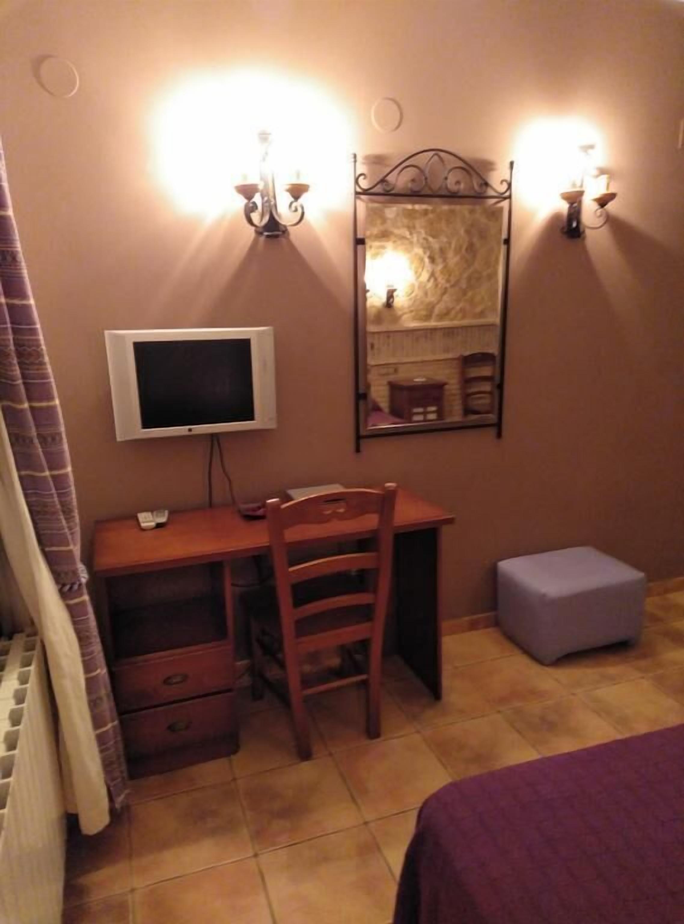 Quarto com escrivaninha de madeira, televisão, cadeira, banquinho pequeno e cama com colcha roxa.