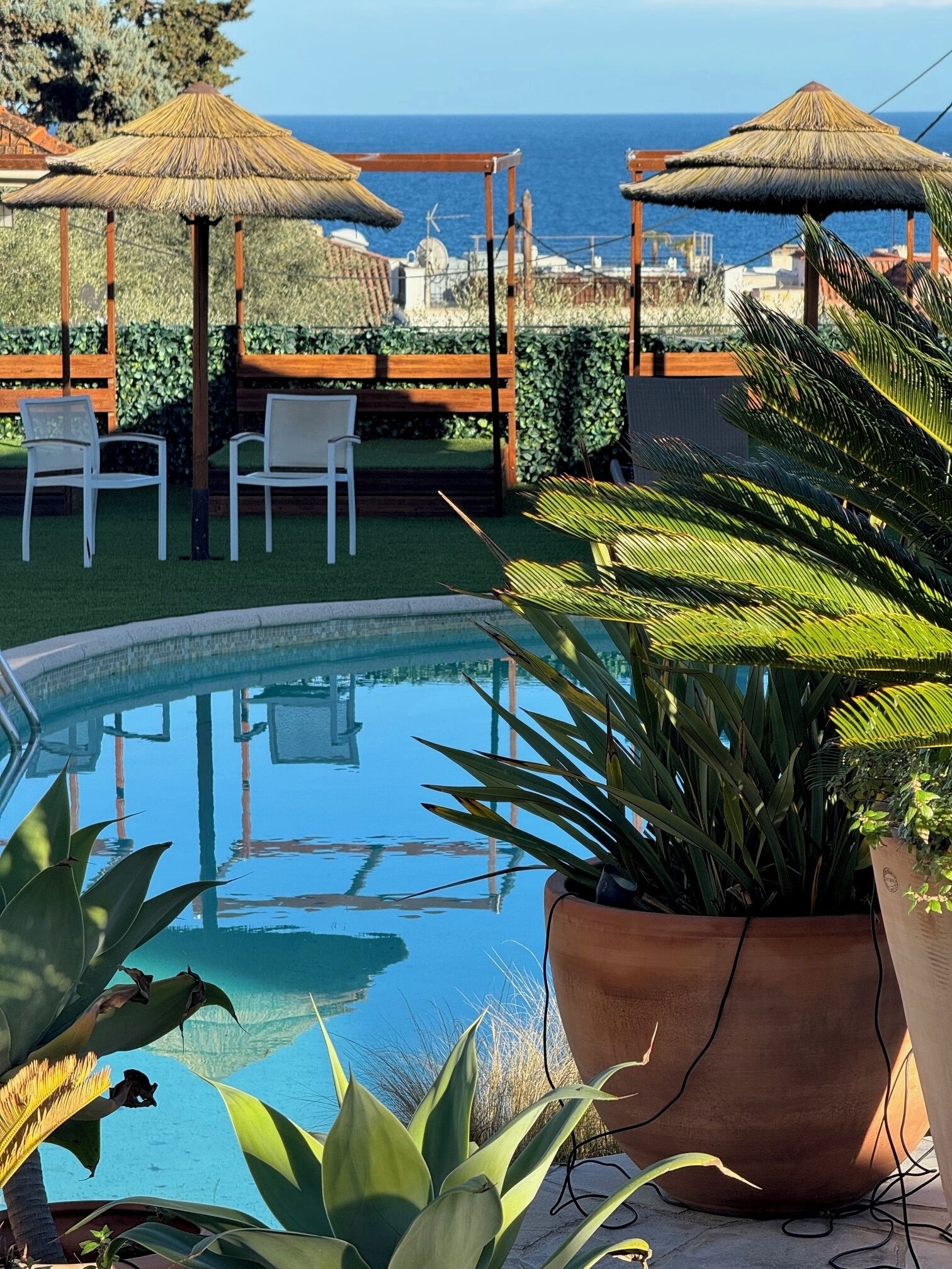 Área da piscina com vista para o mar, cercada por guarda-sóis de palha, cadeiras e vasos com plantas.