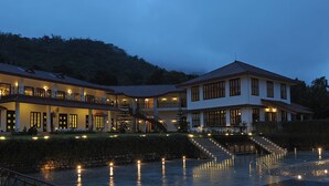 Um edifício grande, de dois andares, com uma piscina em frente, cercado por um jardim paisagístico e uma colina ao fundo.