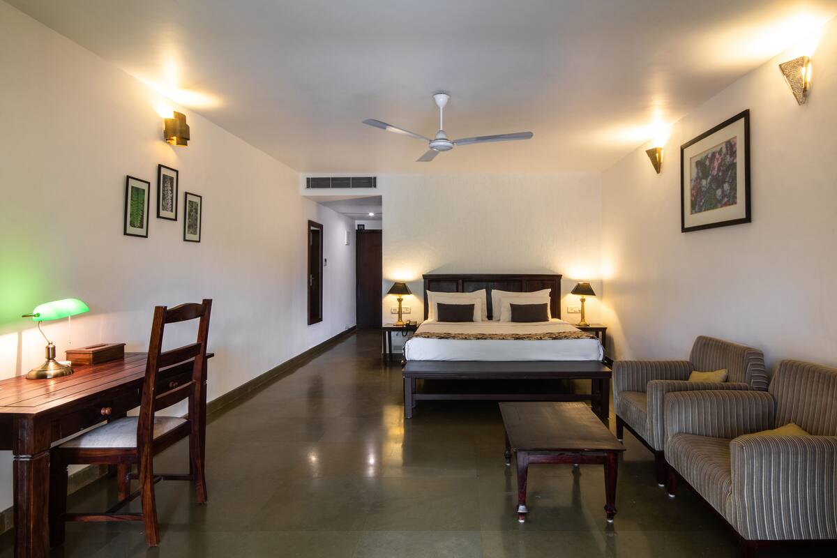 Quarto de hotel com cama, escrivaninha com luminária, poltrona, sofá e ventoinha no teto.