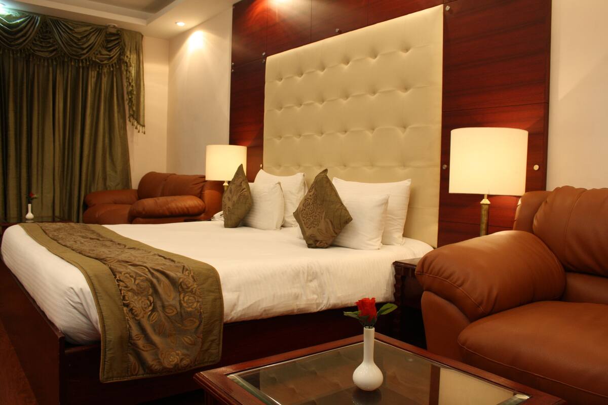 Quarto de hotel com uma cama grande, duas poltronas, uma mesa de centro de vidro e um vaso com uma única rosa.