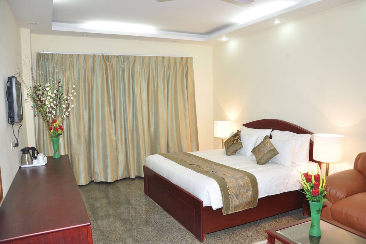 Quarto de hotel com uma cama, duas luminárias laterais, um vaso verde com flores, uma escrivaninha de madeira e uma televisão na parede esquerda.