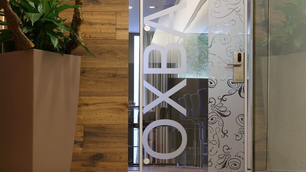 Uma área de recepção com parede de madeira, porta de vidro e placa com a inscrição 'OXBAR', além de um vaso com planta.