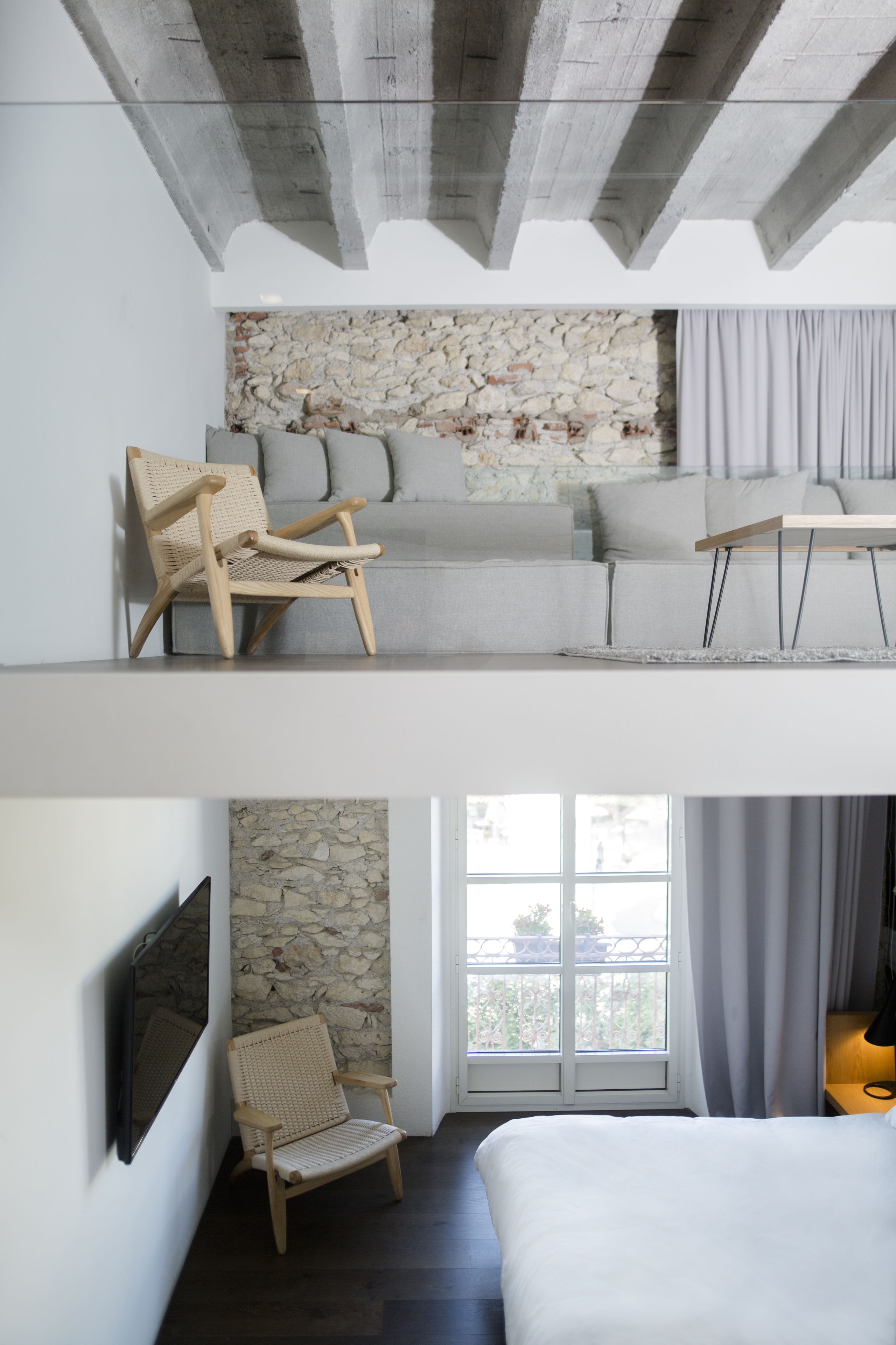 Quarto em dois níveis com parede de pedra, uma cadeira de madeira, sofá, cama e janela com cortinas.