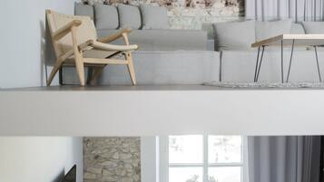 Quarto em dois níveis com parede de pedra, uma cadeira de madeira, sofá, cama e janela com cortinas.