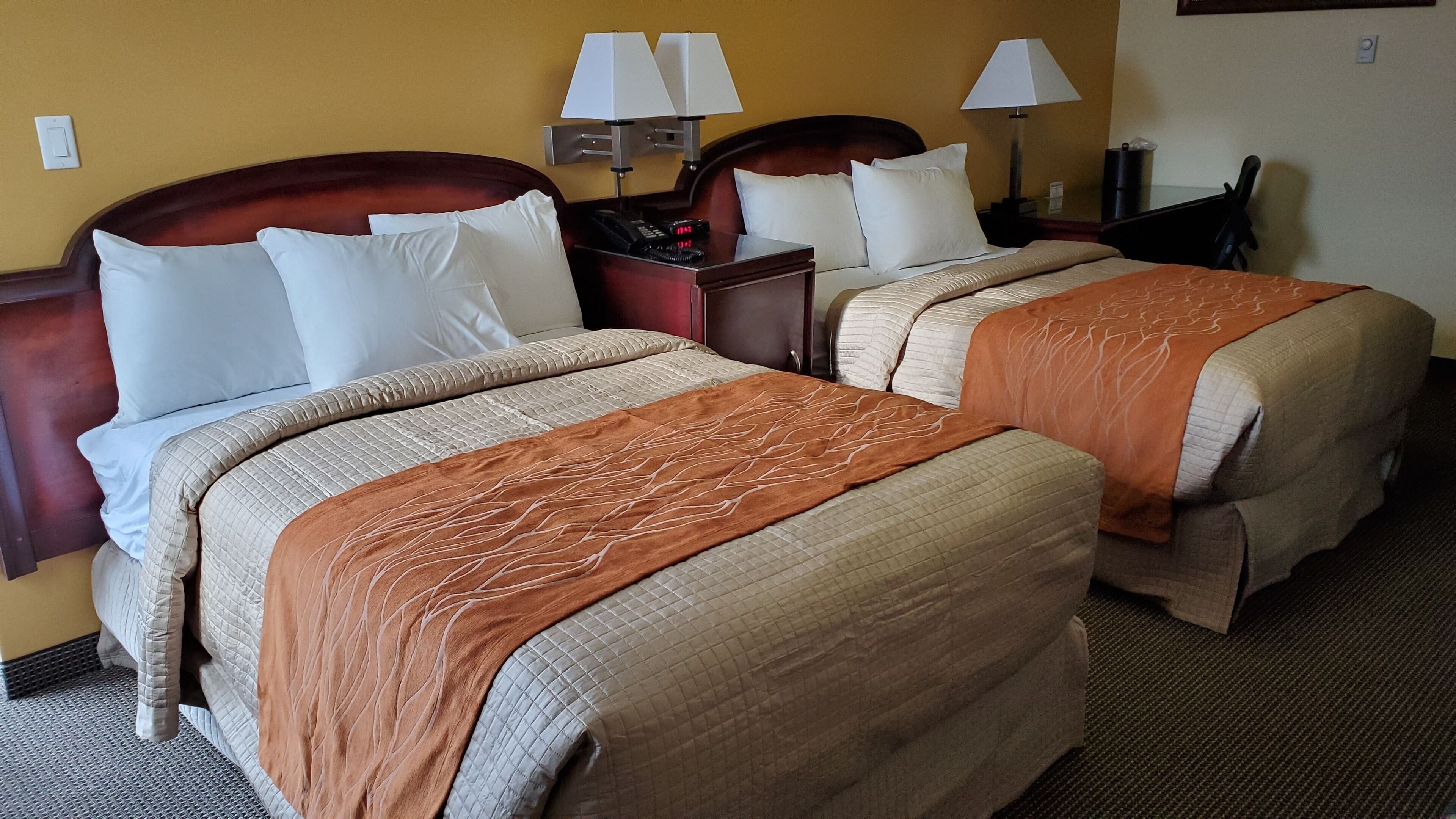 Quarto de hotel com duas camas, cada uma com cabeceira de madeira, roupa de cama branca e manta marrom. Há luminárias de cabeceira, uma mesa de cabeceira com telefone e uma luminária na parede.