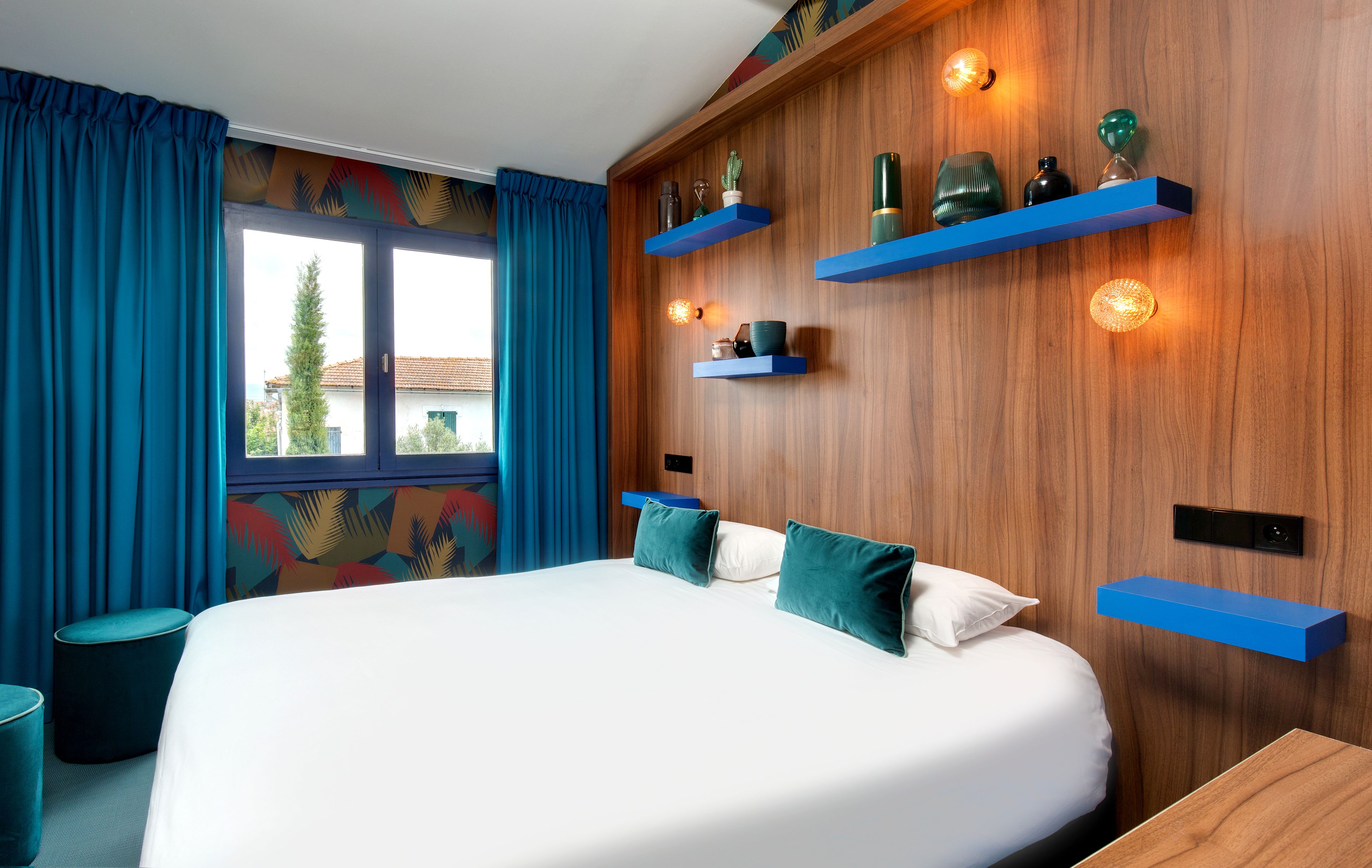 Quarto moderno com uma cama grande, almofadas azuis decorativas e prateleiras de madeira com objetos decorativos.