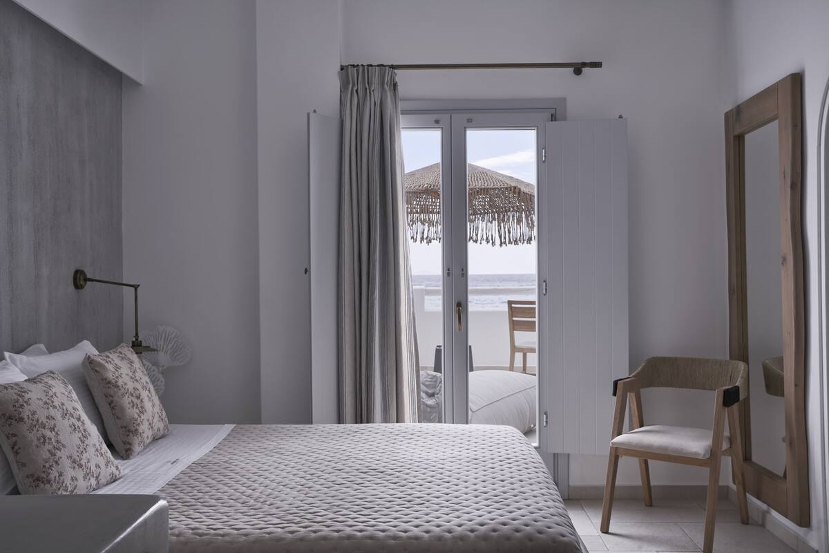 Quarto com cama, uma cadeira e vista para a praia por uma porta aberta.
