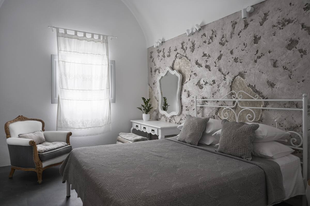 Quarto com cama grande, uma cadeira, cômoda, espelho e janela com cortinas.