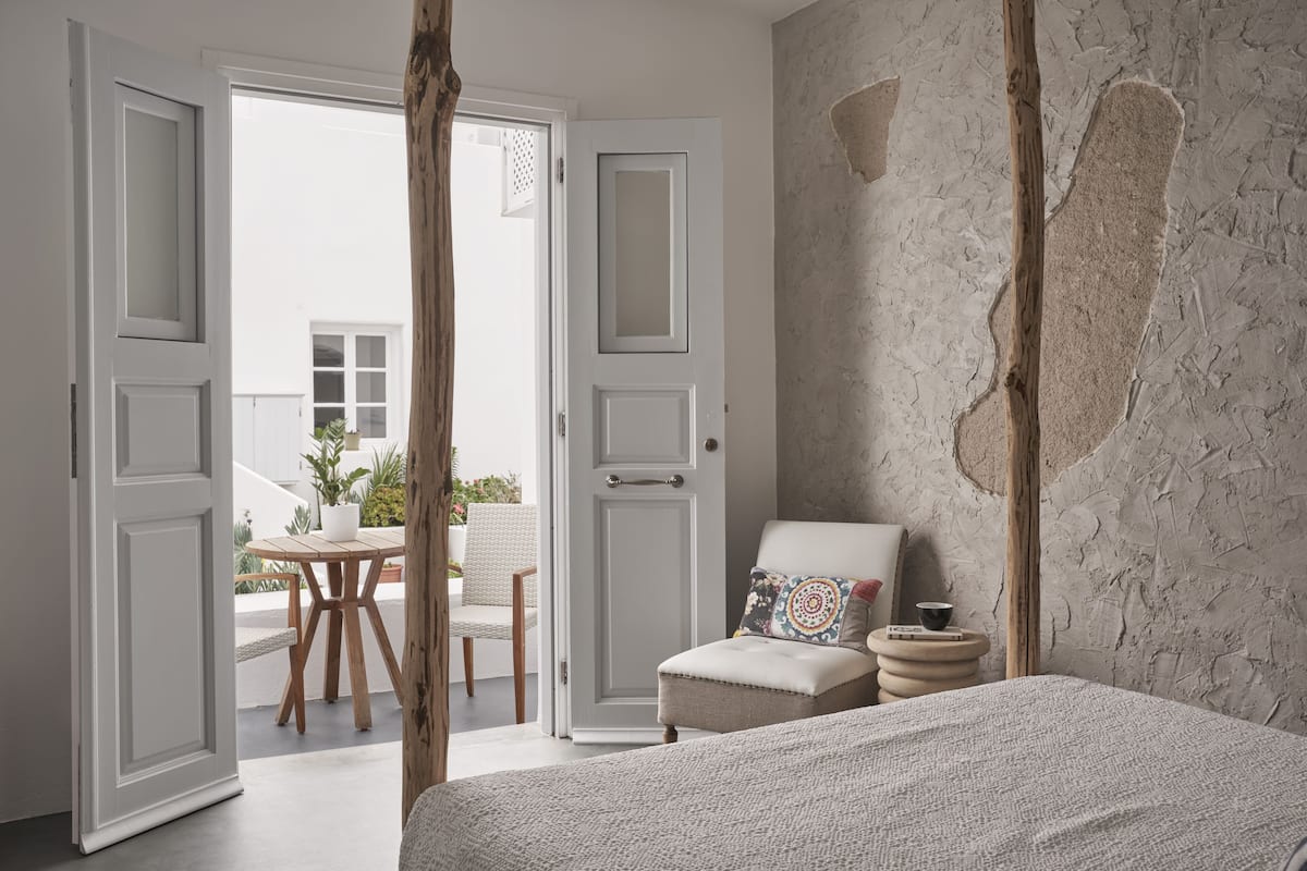 Quarto com uma cama grande, uma cadeira de madeira, uma mesa pequena com uma tigela e vista para a varanda.