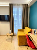 Quarto moderno com sofá amarelo, uma pequena mesa de madeira, cadeira vermelha, televisão de tela plana na parede e uma janela com cortinas.