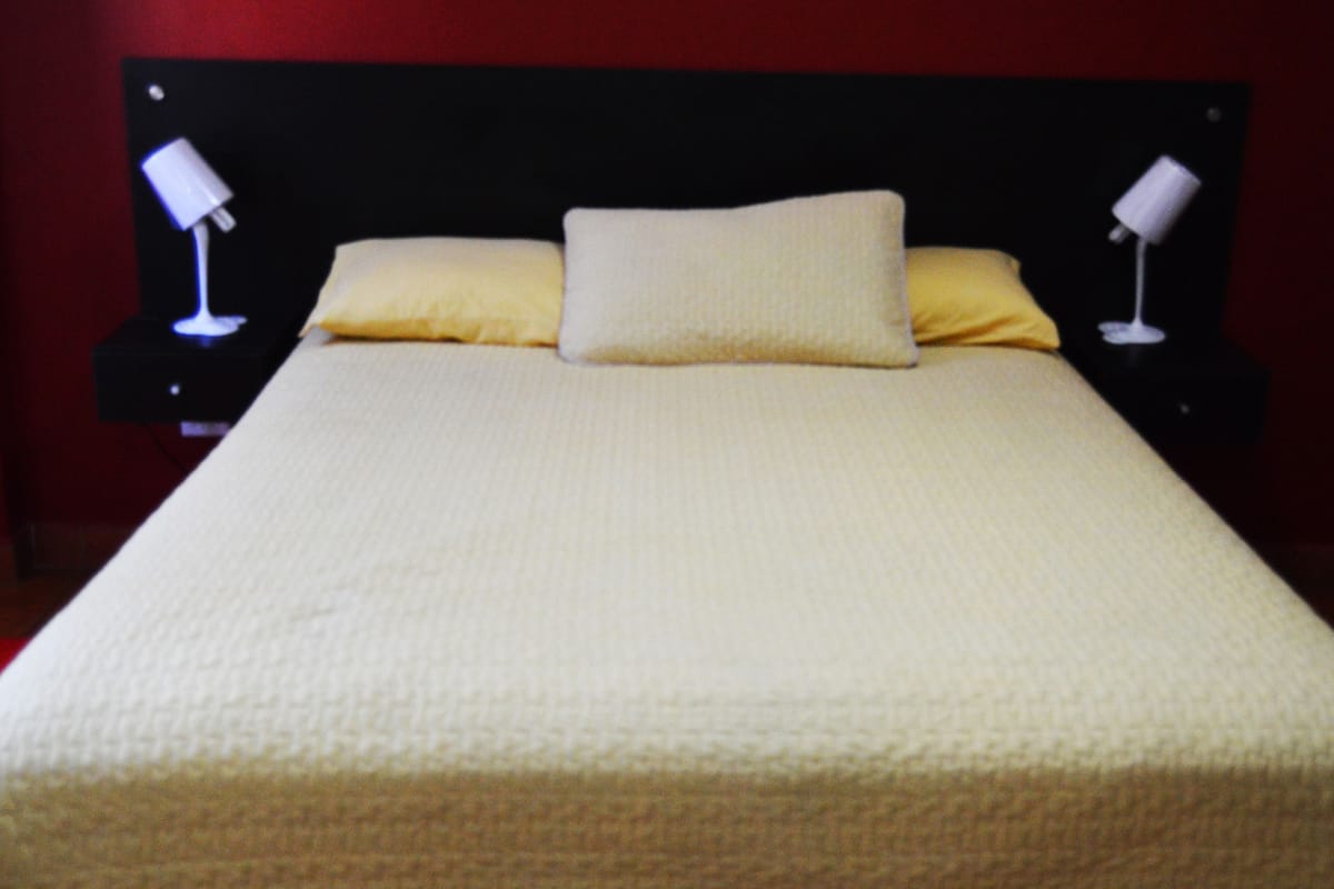 Uma cama com colchão branco e duas almofadas, com duas mesas de cabeceira e abajures, em frente a uma parede vermelha.