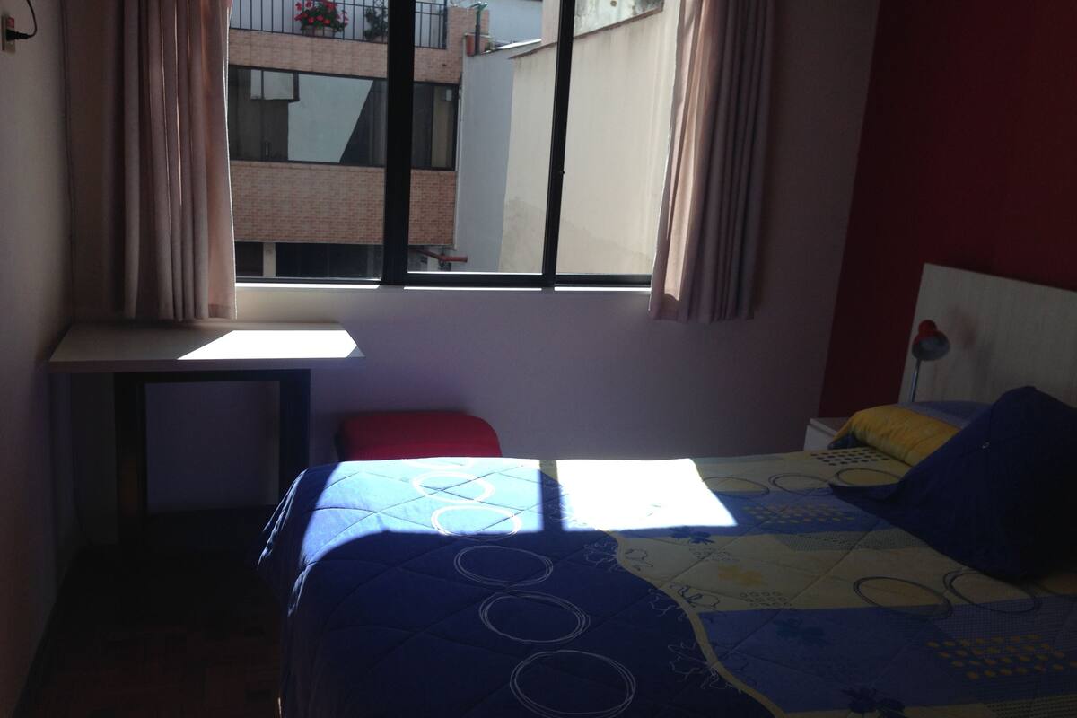 Quarto com cama, escrivaninha e janela.
