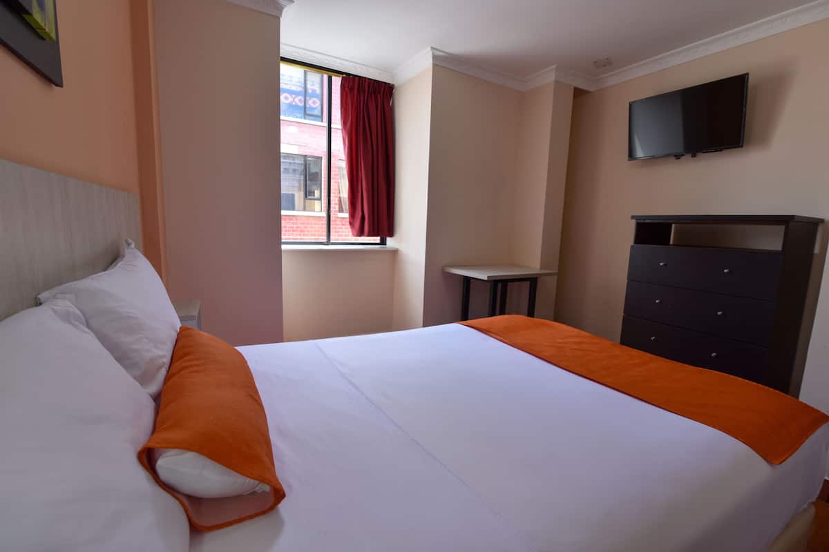 Quarto de hotel com cama, televisão, penteadeira e janela com cortinas.
