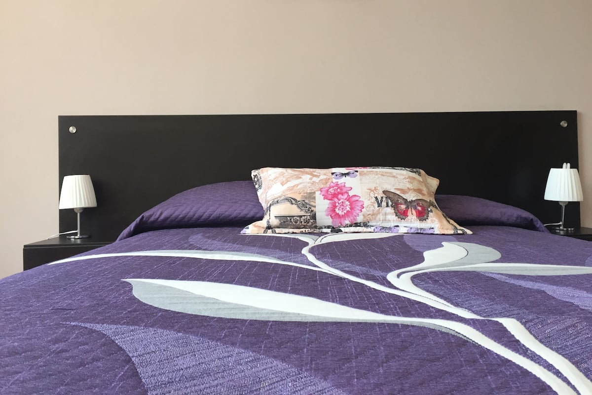 Uma cama com colcha roxa e almofada florida, com duas mesas de cabeceira e abajures de cada lado.