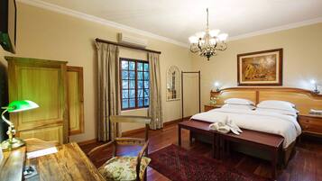 Quarto de hotel com escrivaninha de madeira, uma cadeira, cama com roupa de cama branca, tapete vermelho estampado, lustre e um quadro na parede.